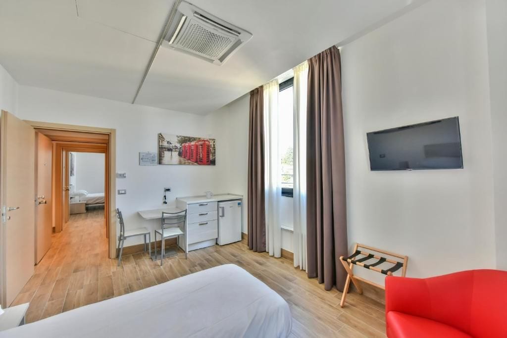 Quarto de hotel com cama, escrivaninha, cadeira, televisão e janela com cortinas.
