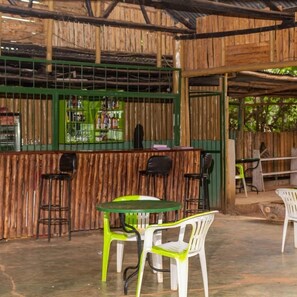 Área de bar ao ar livre com estrutura de madeira, balcão verde e cadeiras de plástico branco.