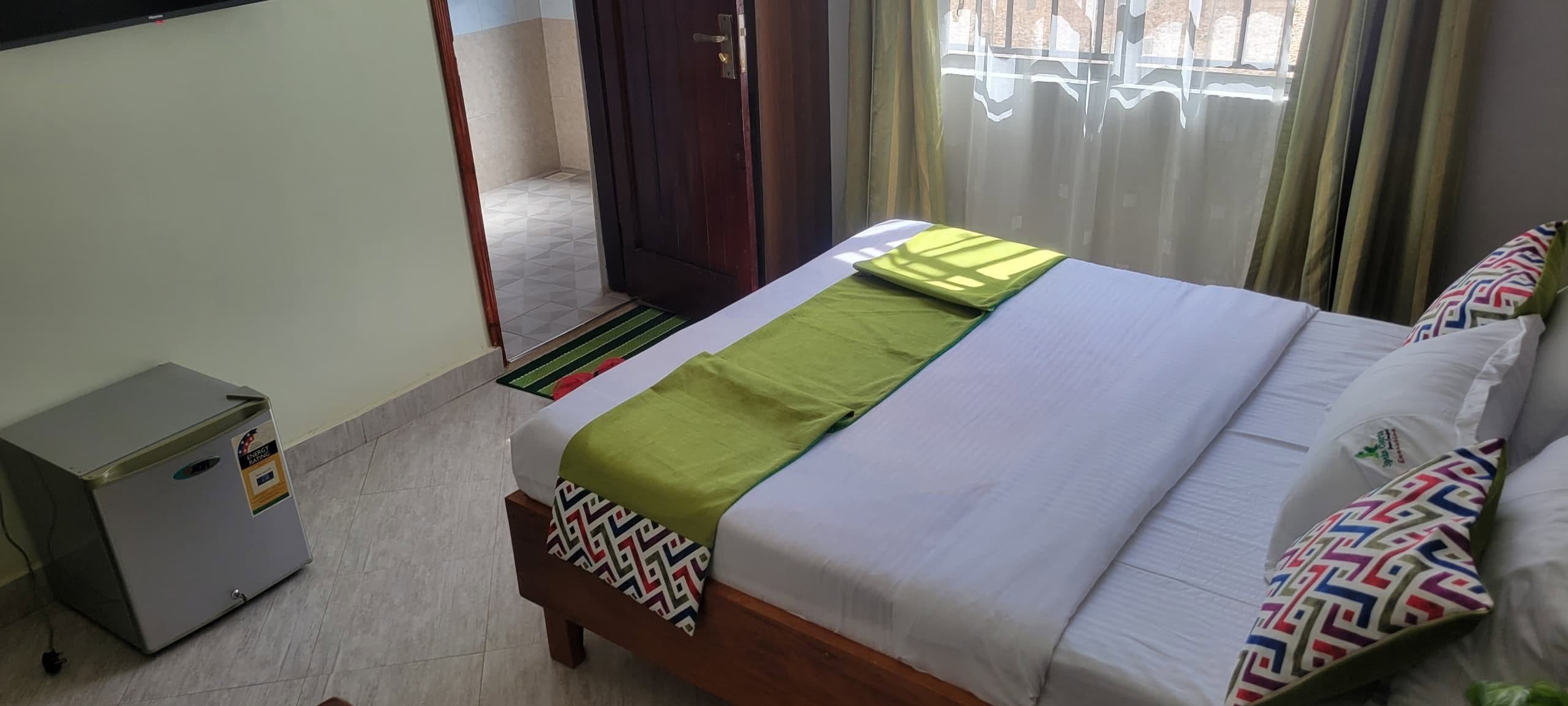 Quarto de hotel com cama, pequeno frigorífico, porta e janela com cortinas.