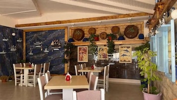 Interior de restaurante com mesas e cadeiras de madeira, um mural na parede e plantas em vasos.