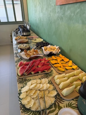 Mesa de buffet com diversas opções de comida, incluindo frutas, doces e uma máquina de café.