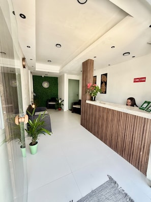 Uma área de recepção moderna com um balcão de madeira, uma parede verde com relógio e plantas em vasos.