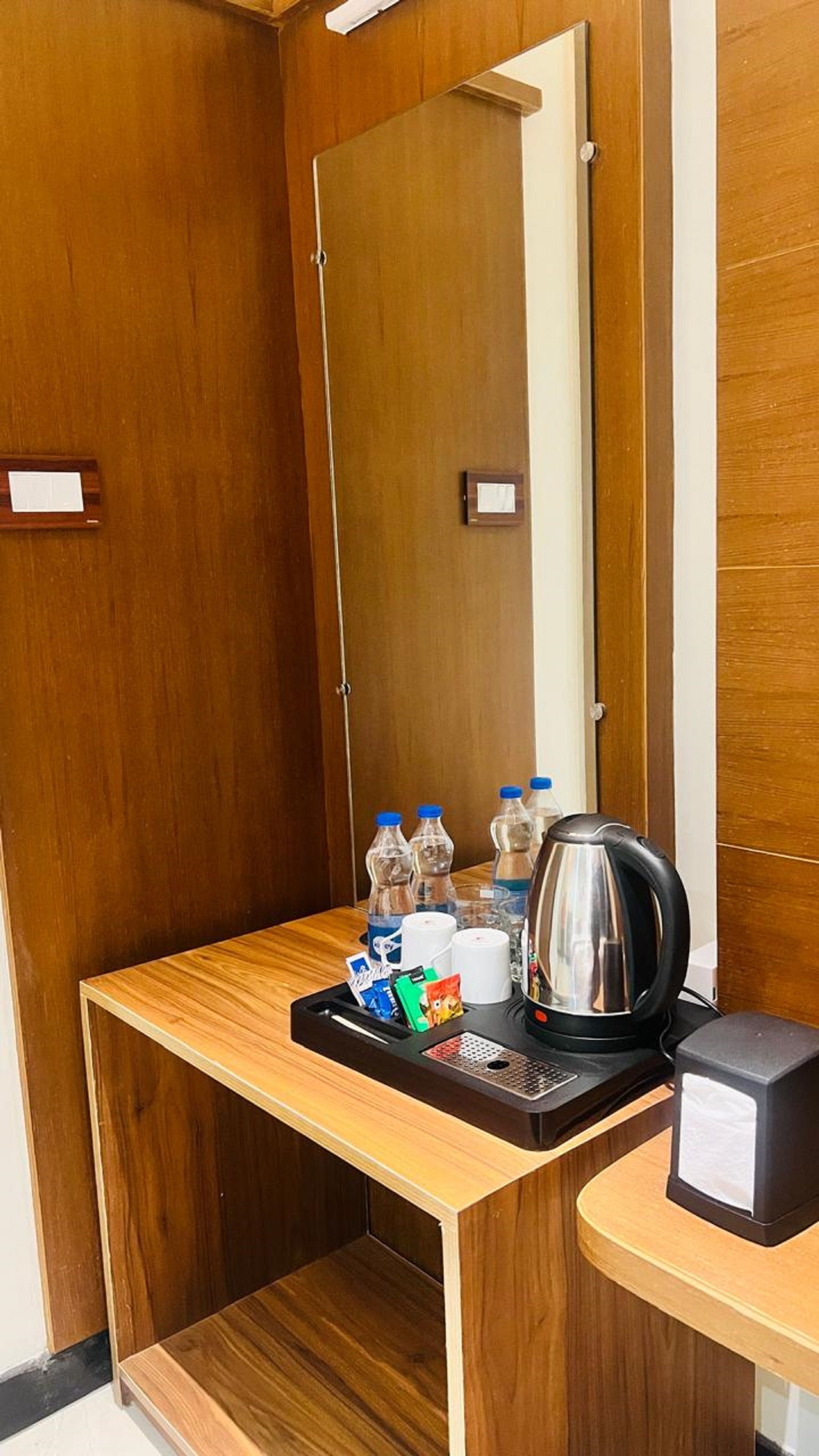 Quarto de hotel com escrivaninha de madeira, chaleira, água engarrafada e bandeja com diversos objetos.