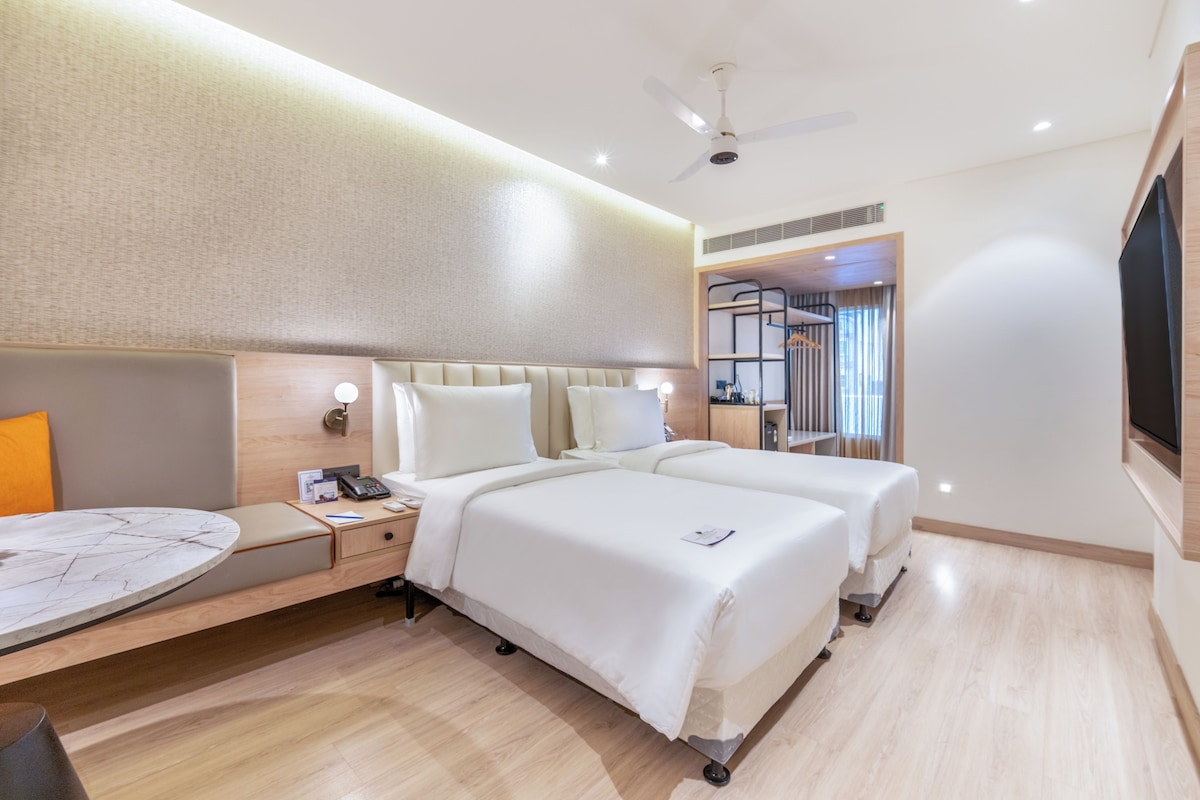 Quarto de hotel com duas camas, uma escrivaninha, uma cadeira, uma mesa redonda, um ventilador de teto e uma televisão.