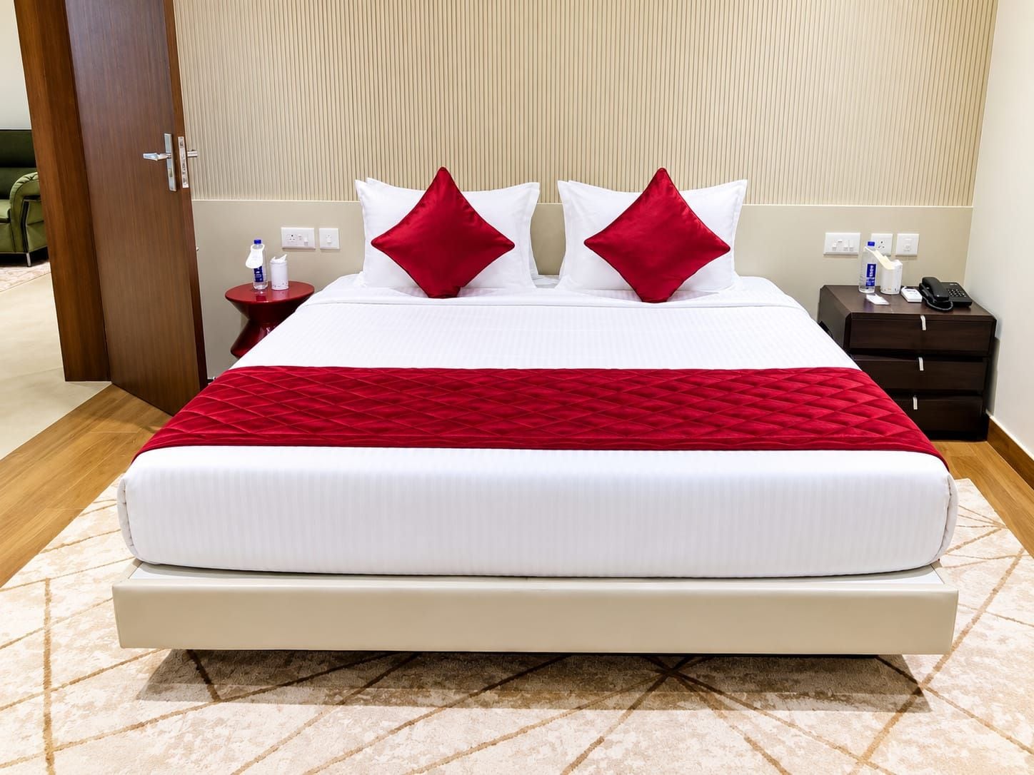Quarto de hotel com uma cama grande, um cobertor vermelho e uma mesa de cabeceira com um telefone e uma garrafa.