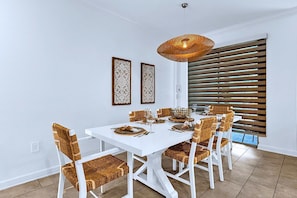 Uma área de refeições com mesa branca, cadeiras de vime e luminária pendente trançada.
