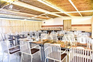 Ampla sala de jantar com várias mesas e cadeiras, teto alto com telhado de palha e um serviço de buffet.