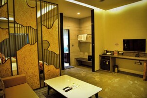 Quarto de hotel com mesa branca, televisão, cozinha compacta e banheiro visível através de uma porta aberta.