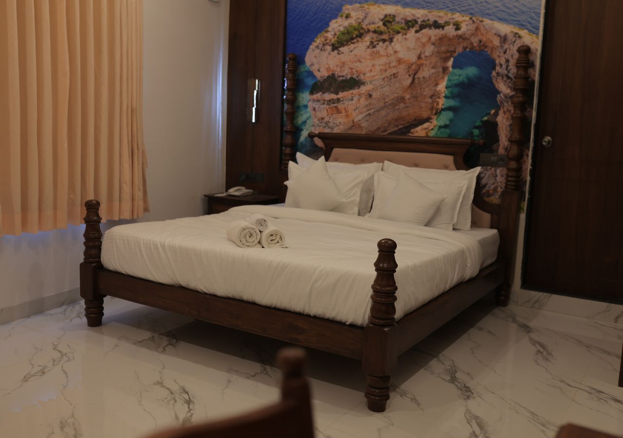 Cama bem arrumada com estrutura de madeira e roupa de cama branca, mesa de cabeceira com abajur e uma grande pintura de paisagem costeira na parede.