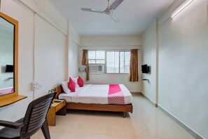 Quarto de hotel com cama, escrivaninha, cadeira, ventilador de teto, ar condicionado e janela com cortinas.