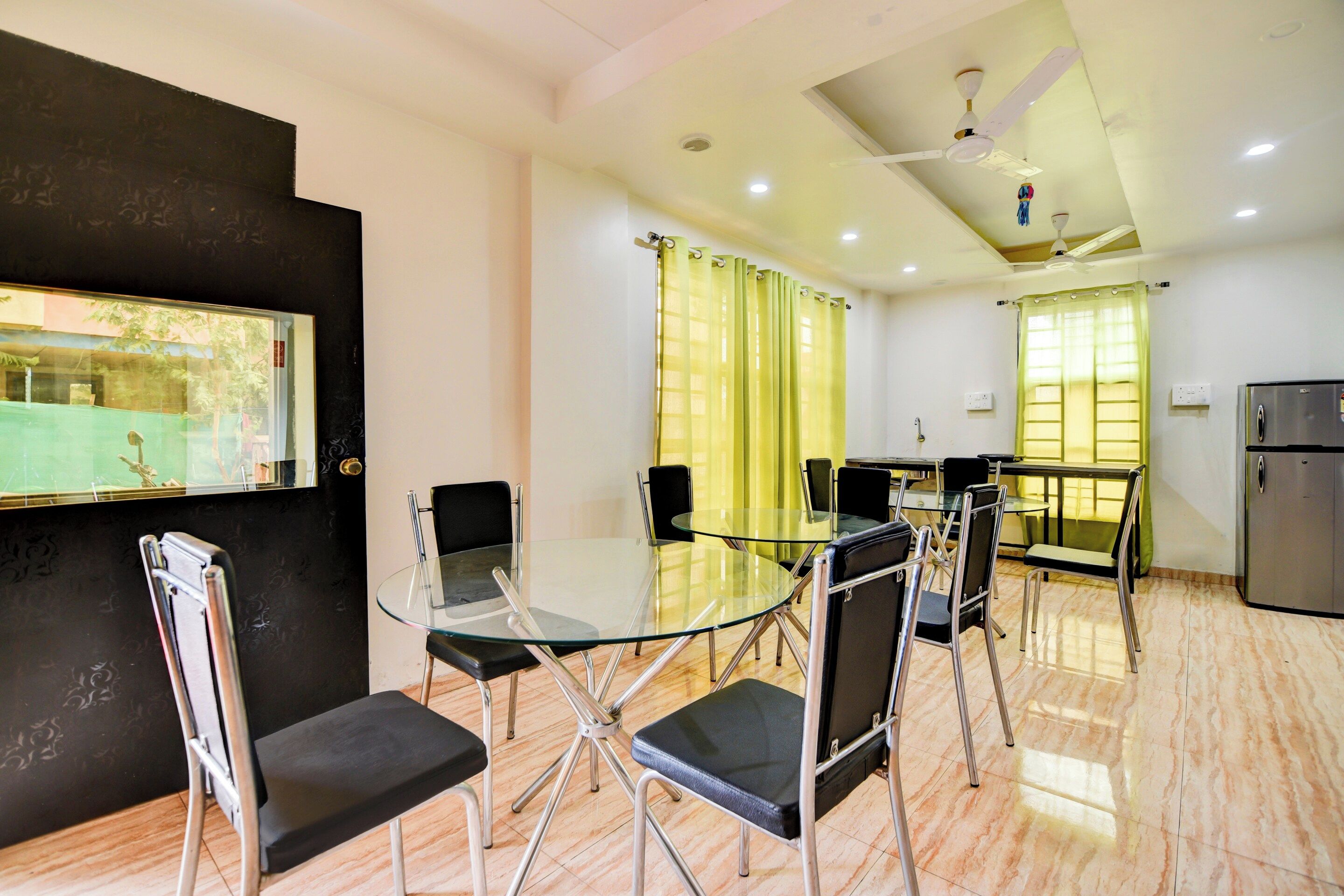 Uma área de refeições moderna com mesa de vidro e cadeiras de metal, cozinha com geladeira e ventilador de teto.