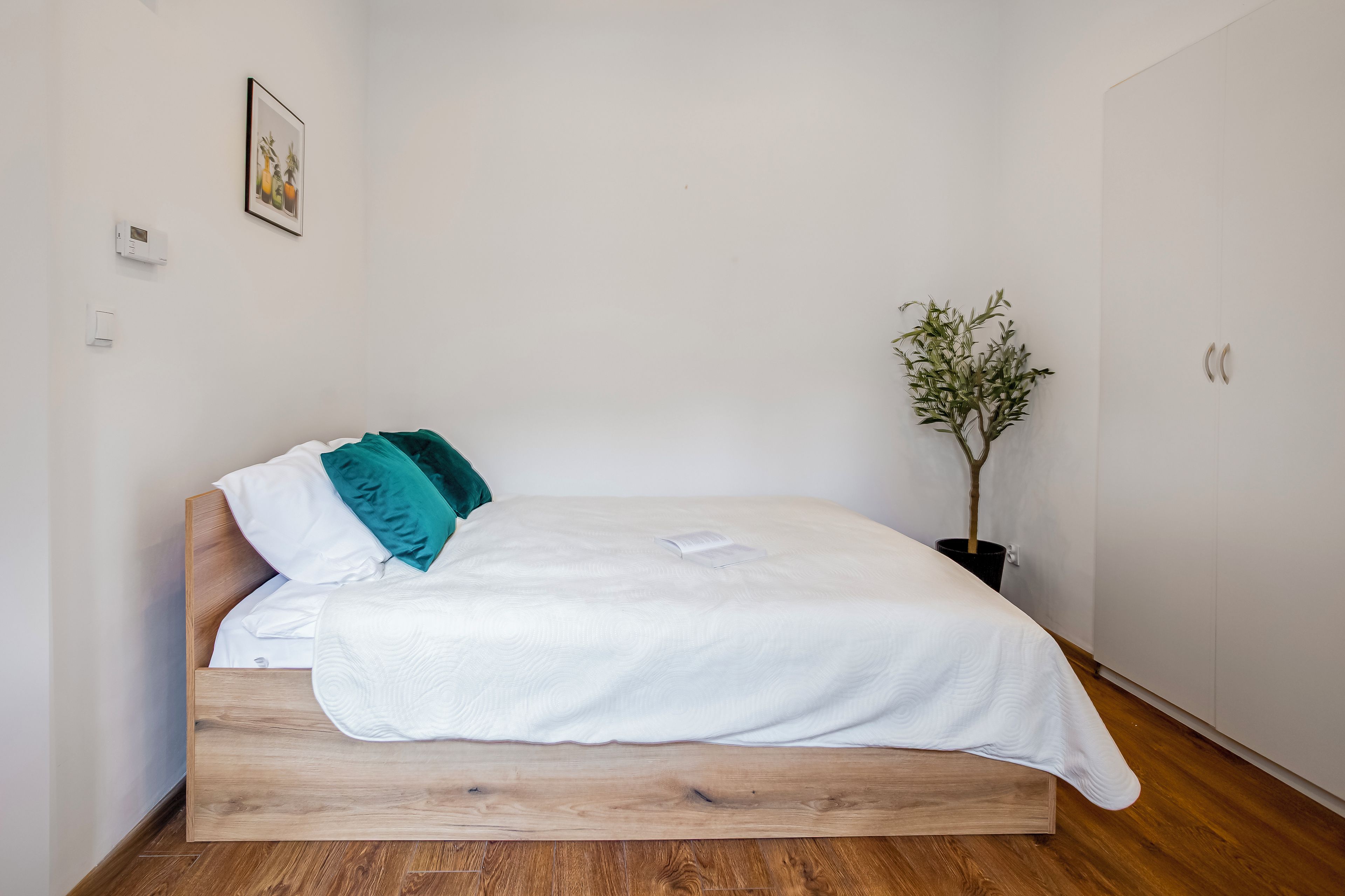 Uma cama de solteiro com estrutura de madeira e roupa de cama branca, uma pequena planta em vaso e um termostato fixado na parede.