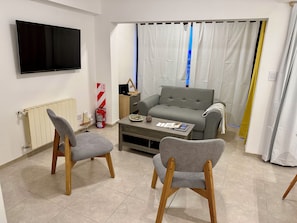 Uma sala de estar com sofá cinza, duas poltronas cinzas, mesa de centro, televisão de tela plana, extintor de incêndio e janela com cortinas.