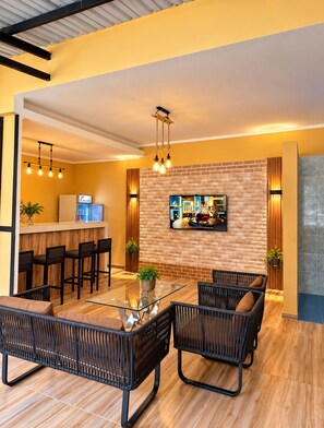 Uma sala de estar moderna com uma mesa de centro de vidro, móveis de vime, uma TV de tela plana presa a uma parede de tijolos e uma área de cozinha com banquetas.