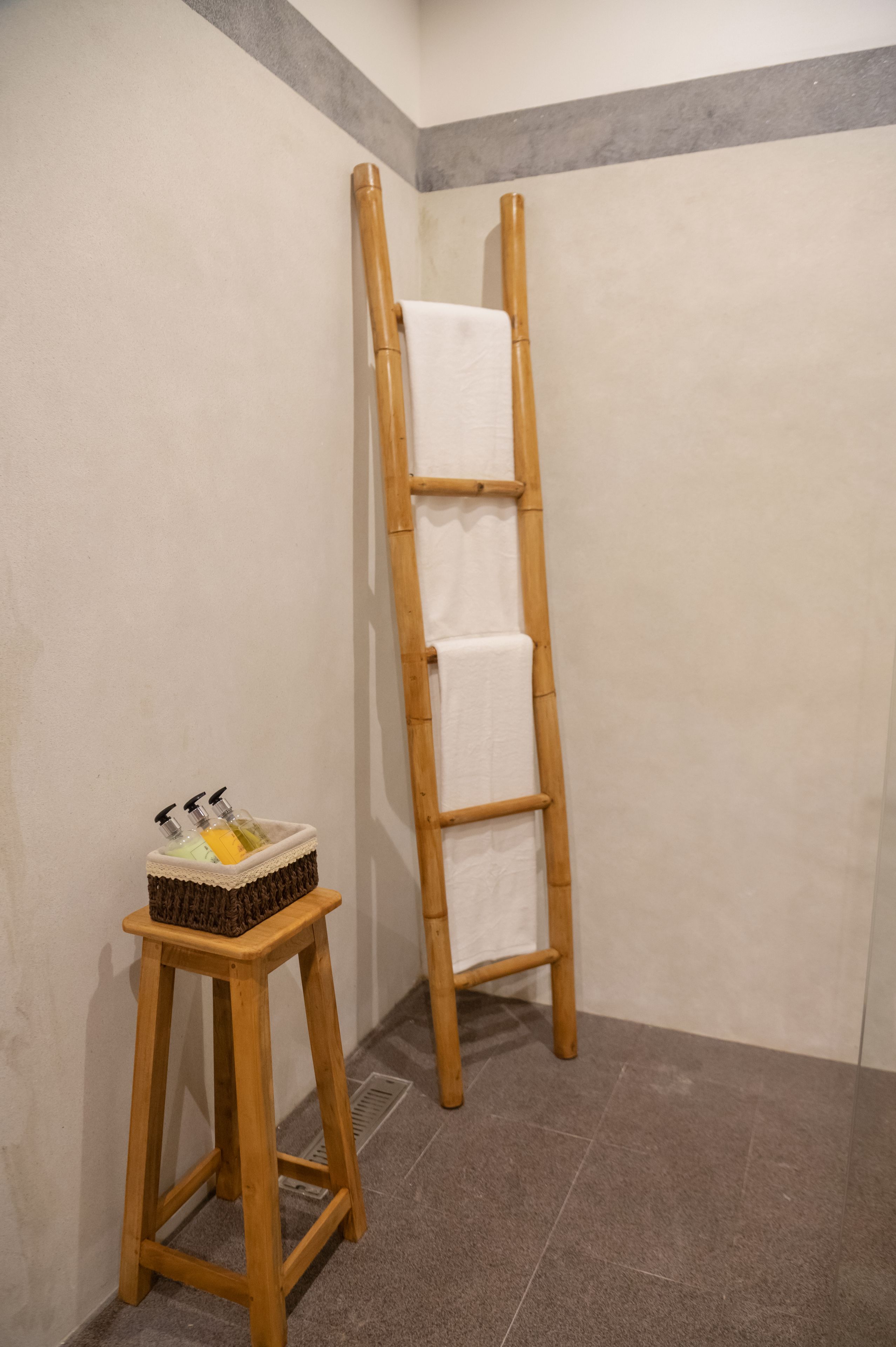 Canto de um quarto com uma escada de madeira encostada na parede, um banquinho de madeira e uma cesta de produtos de higiene pessoal sobre ele, e piso de azulejos.