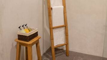 Canto de um quarto com uma escada de madeira encostada na parede, um banquinho de madeira e uma cesta de produtos de higiene pessoal sobre ele, e piso de azulejos.