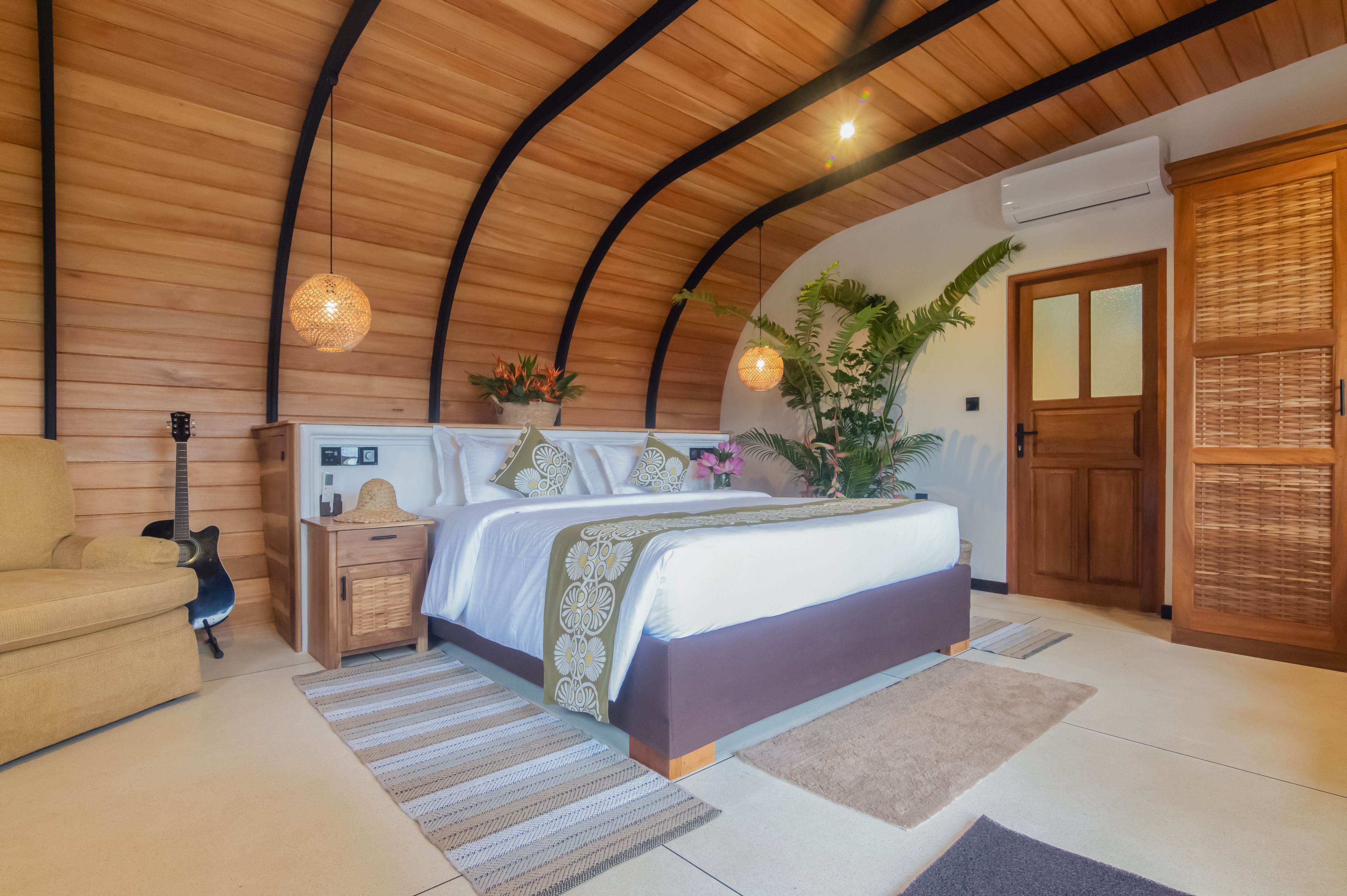 Quarto com teto de madeira curvo, cama com colcha estampada, uma guitarra, mesa de cabeceira e um vaso com planta.