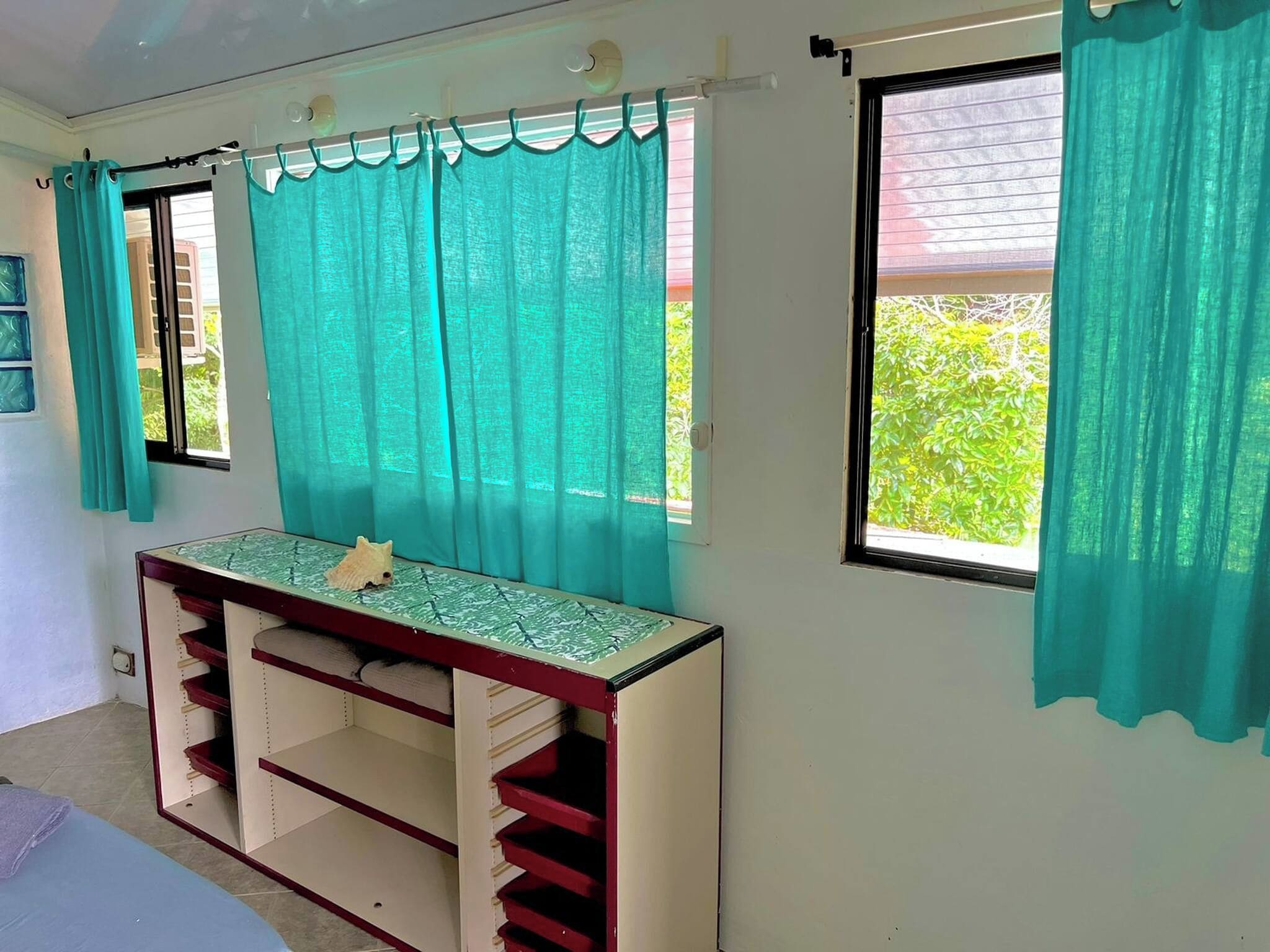 Quarto com escrivaninha, duas janelas com cortinas e vista para o verde lá fora.
