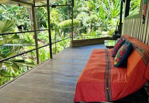 Um deck de madeira com um banco e almofadas coloridas, cercado por folhagem tropical exuberante.