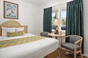 Quarto de hotel com cama, duas cadeiras, mesa com luminária, pintura na parede e janela com cortinas.