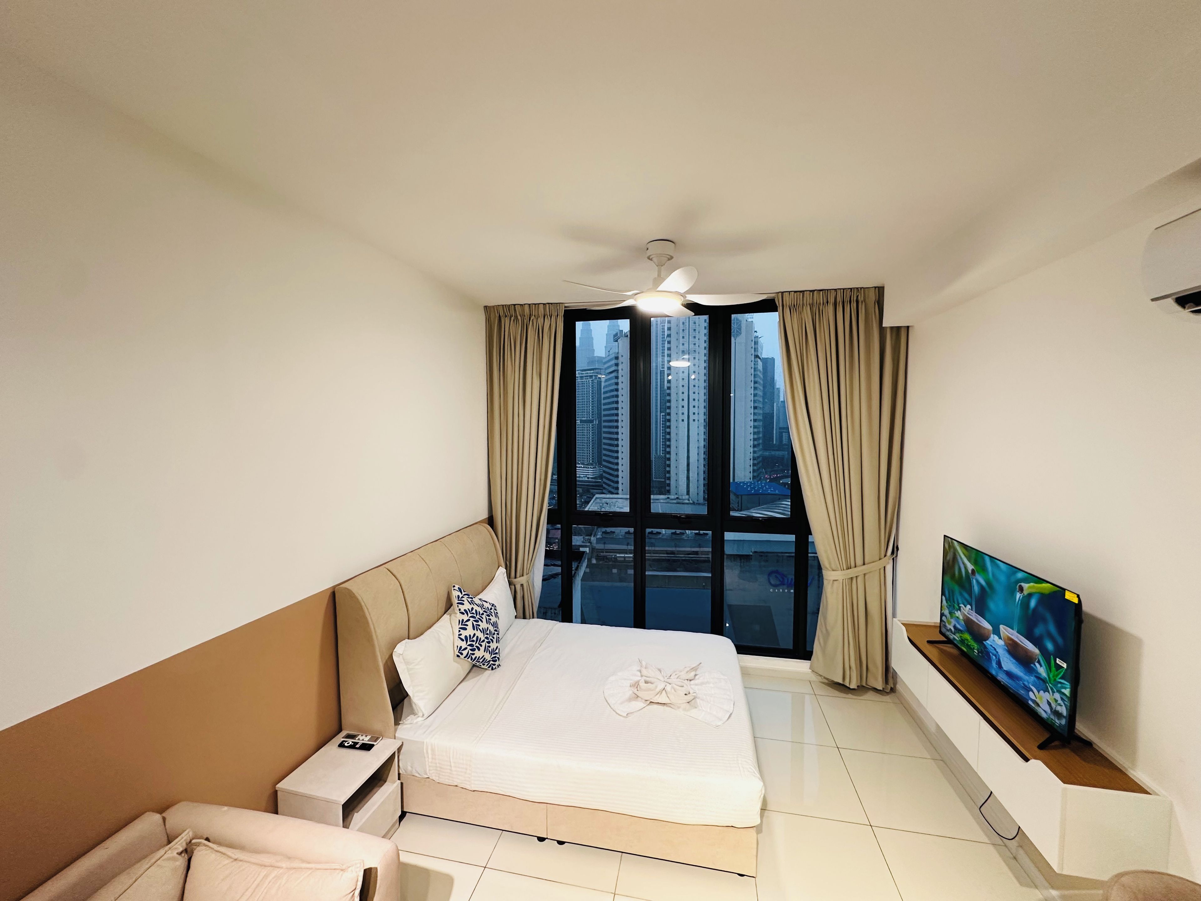 Quarto de hotel com cama, televisão, janela com cortinas e ventoinha no teto.