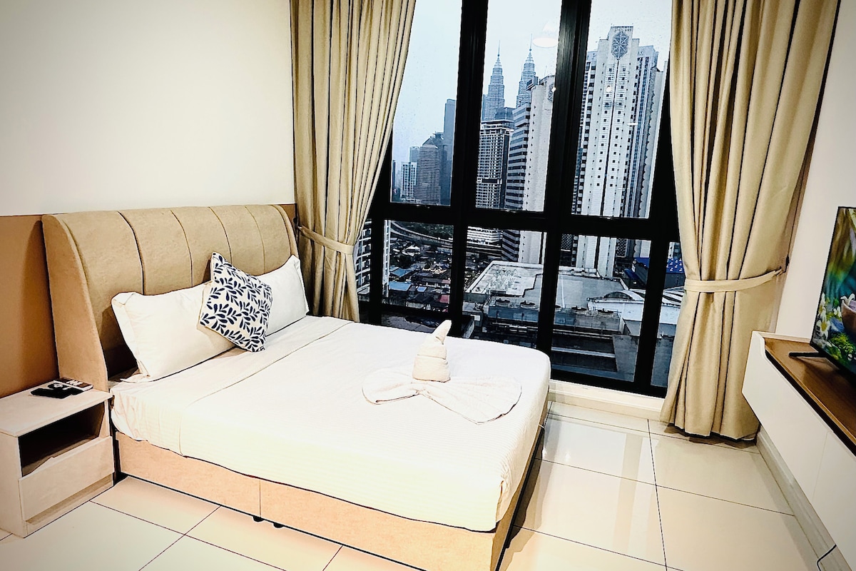 Quarto de hotel com cama, mesa de cabeceira, janela com cortinas e vista para a paisagem urbana.