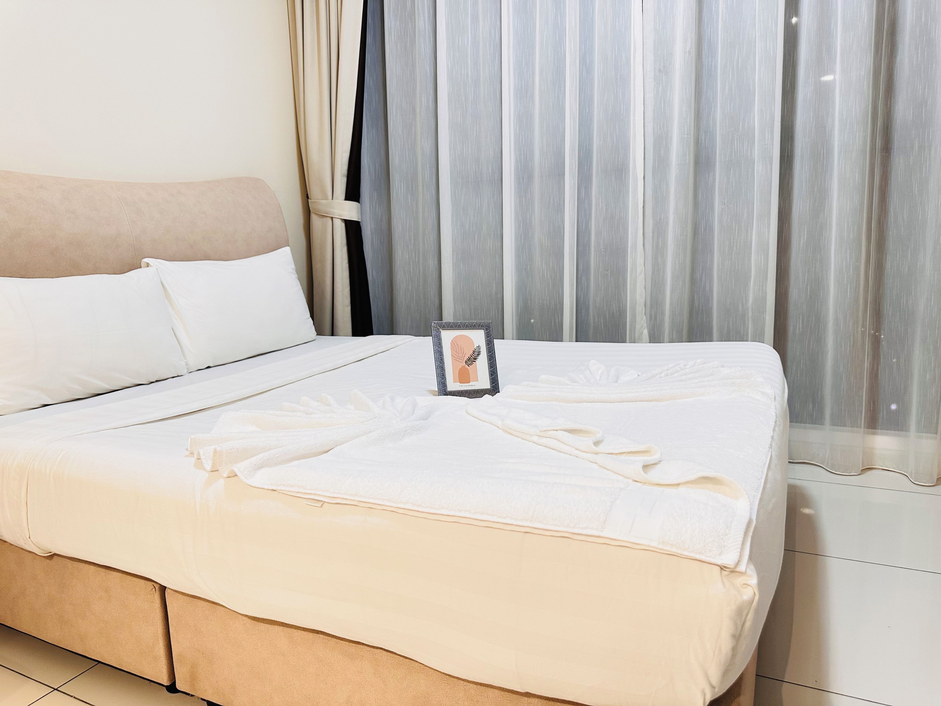 Uma cama bem arrumada com edredom branco e almofadas, um quadro emoldurado sobre a cama e uma janela com cortinas leves.