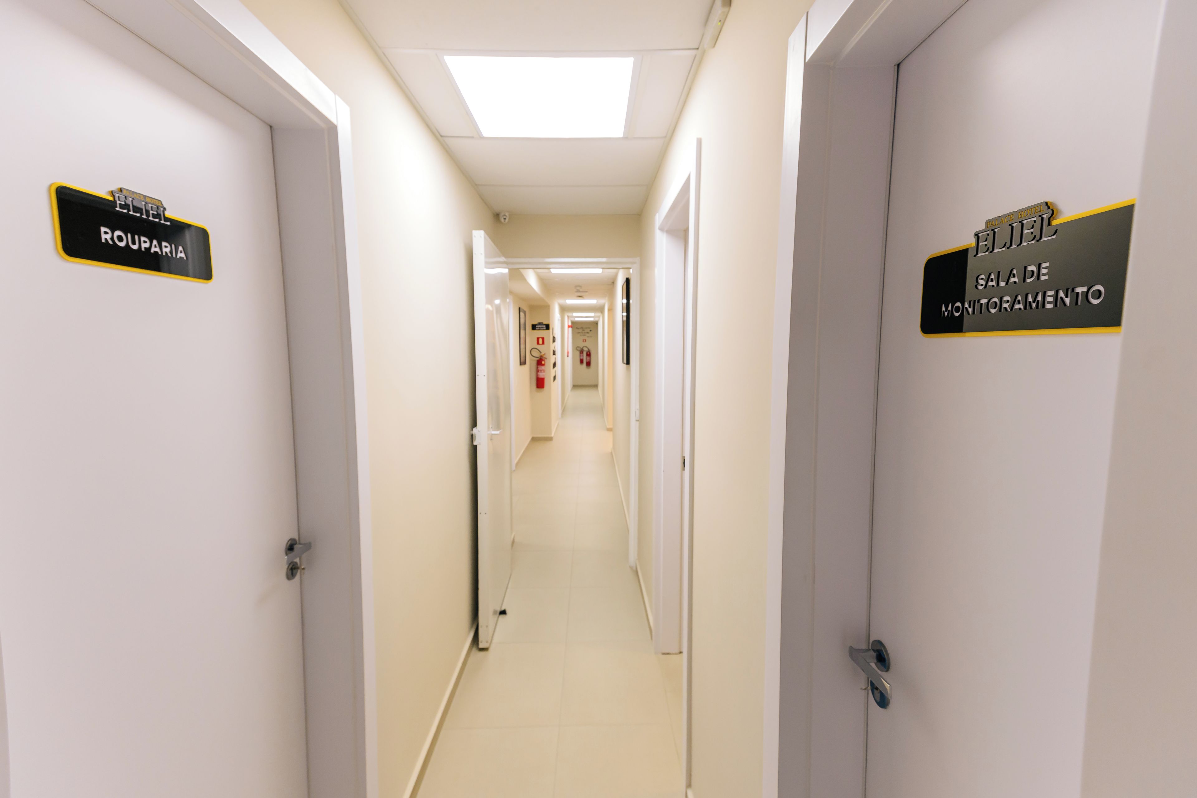 Um corredor de hotel com portas identificadas como 'ELIEL ROUPARIA' e 'ELIEL SALA DE MONITORAMENTO'.