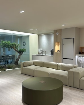 Uma sala de estar moderna com sofá em formato de L, um banquinho redondo, uma parede de vidro com uma árvore bonsai, área de cozinha com geladeira e iluminação embutida no teto.