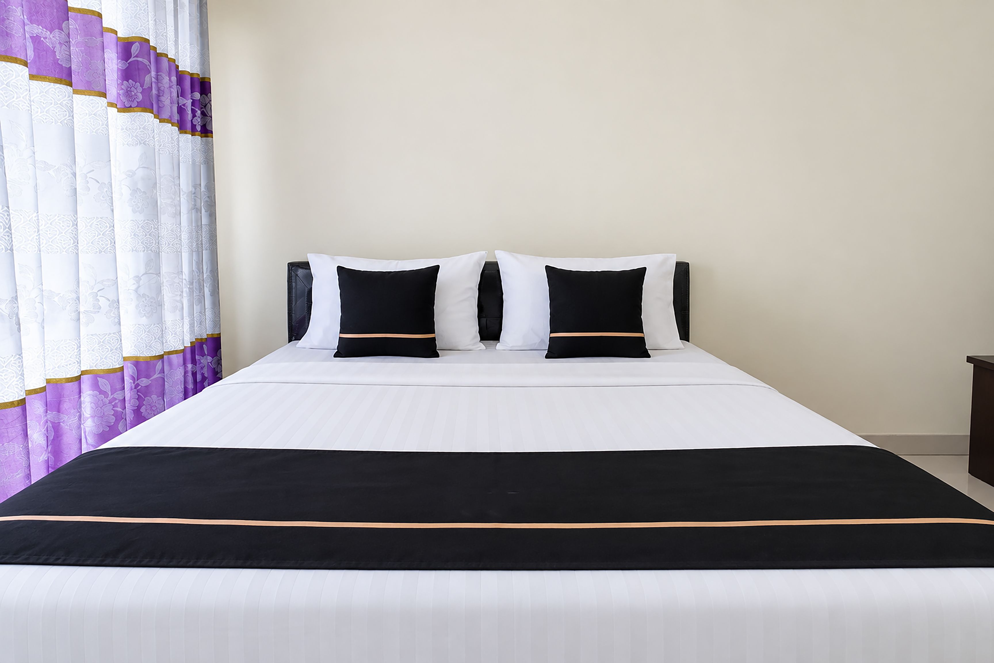 Uma cama com colcha preta e branca, duas almofadas pretas com detalhes dourados e uma cortina roxa e branca.
