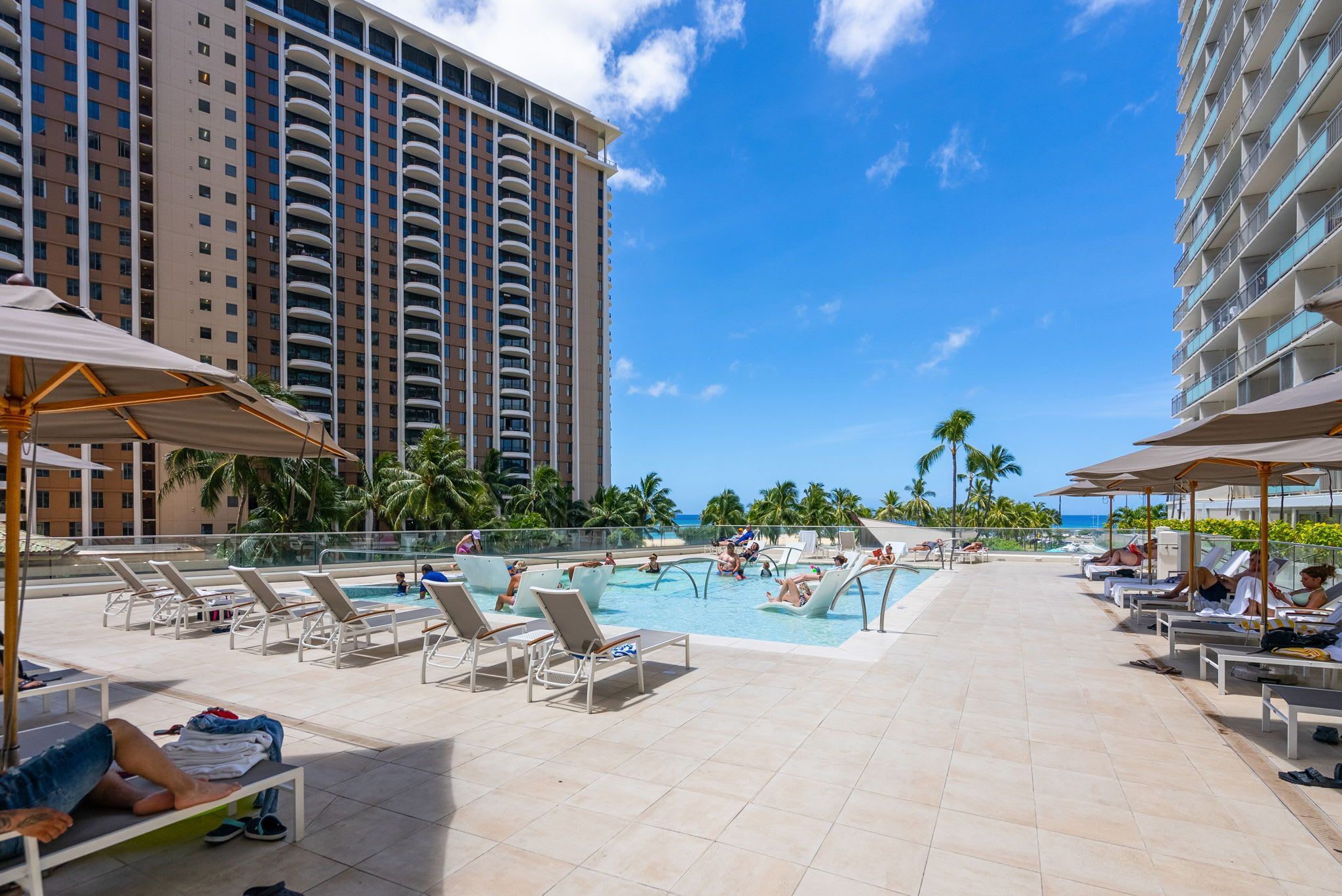 Área de piscina em resort com espreguiçadeiras, piscina e vista para edifícios altos e palmeiras.