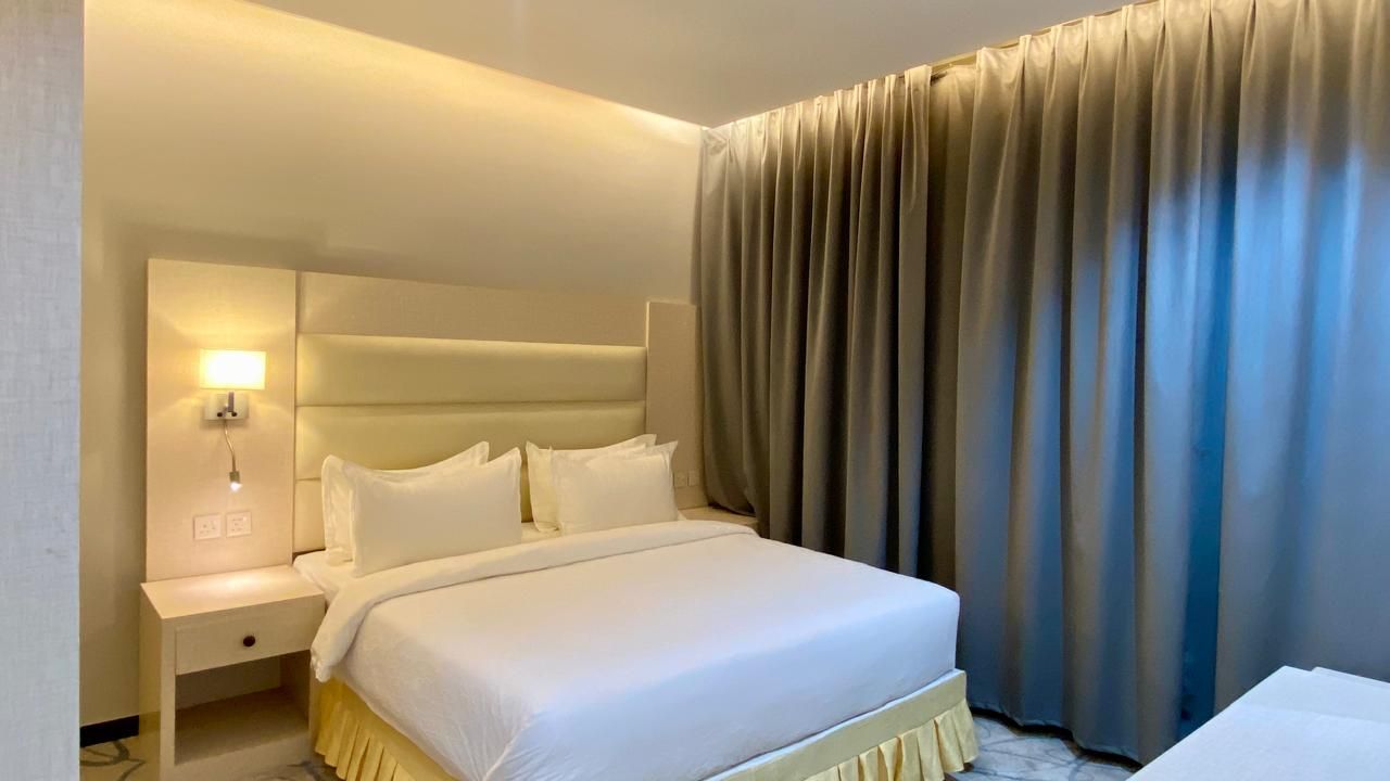 Quarto de hotel com cama, mesa de cabeceira, luminária e janela com cortinas.