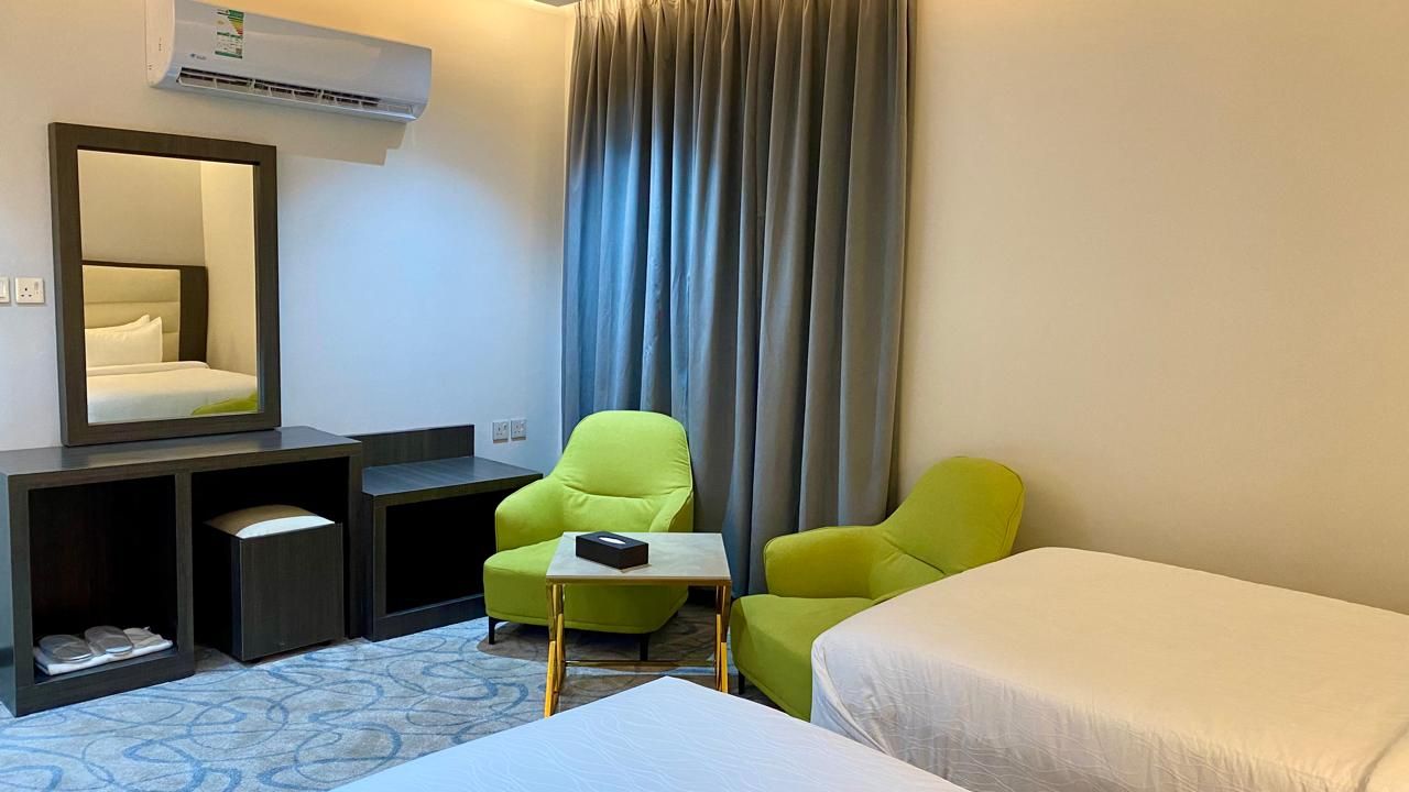 Quarto de hotel com cama, escrivaninha, cadeira, mesinha, espelho e janela com cortinas.