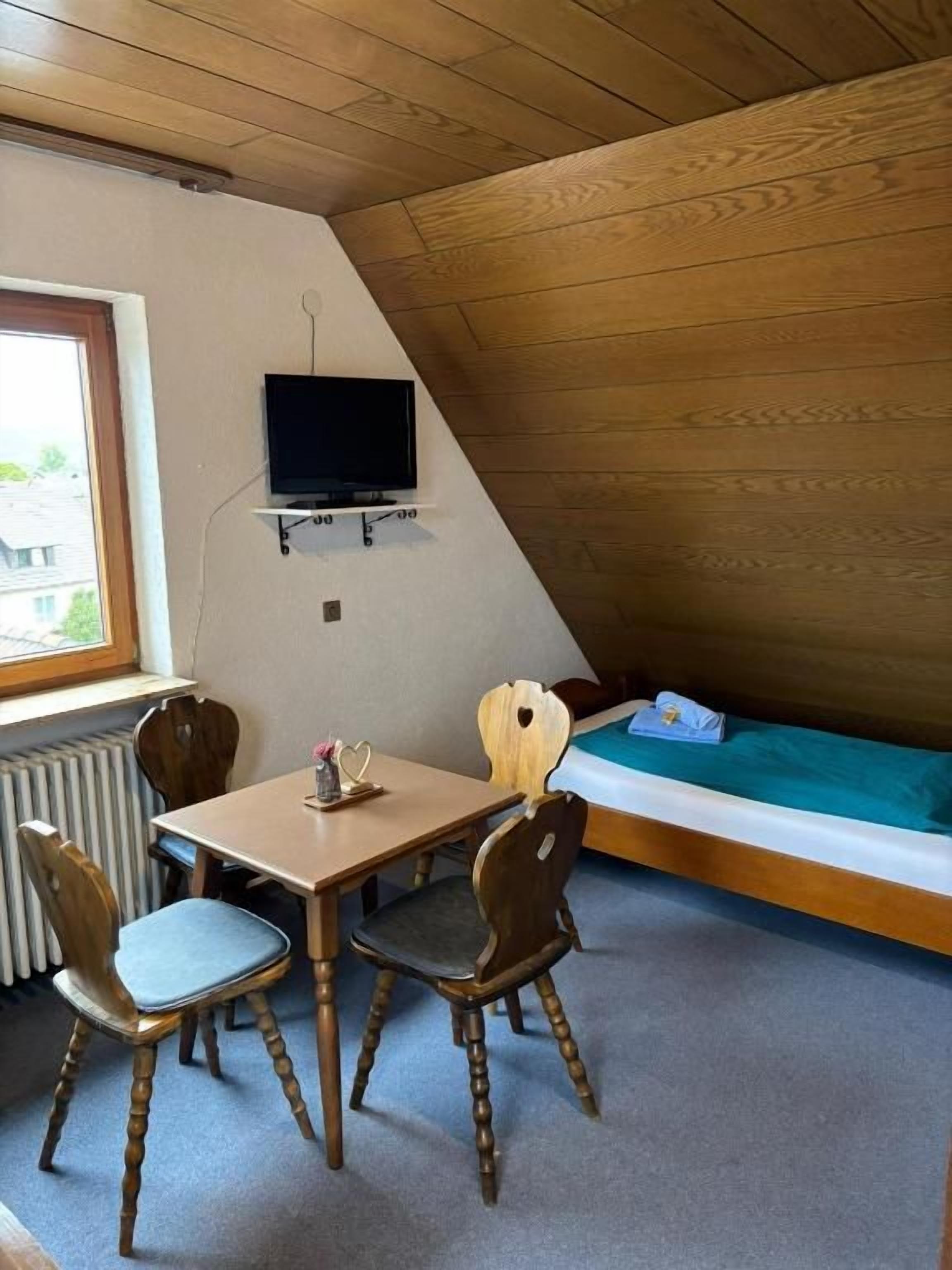 Quarto com teto inclinado, cama com colchão azul, mesa com cadeiras, televisão fixada na parede e janela com vista para o verde.