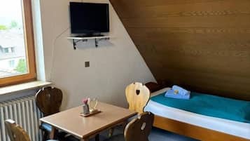 Quarto com teto inclinado, cama com colchão azul, mesa com cadeiras, televisão fixada na parede e janela com vista para o verde.