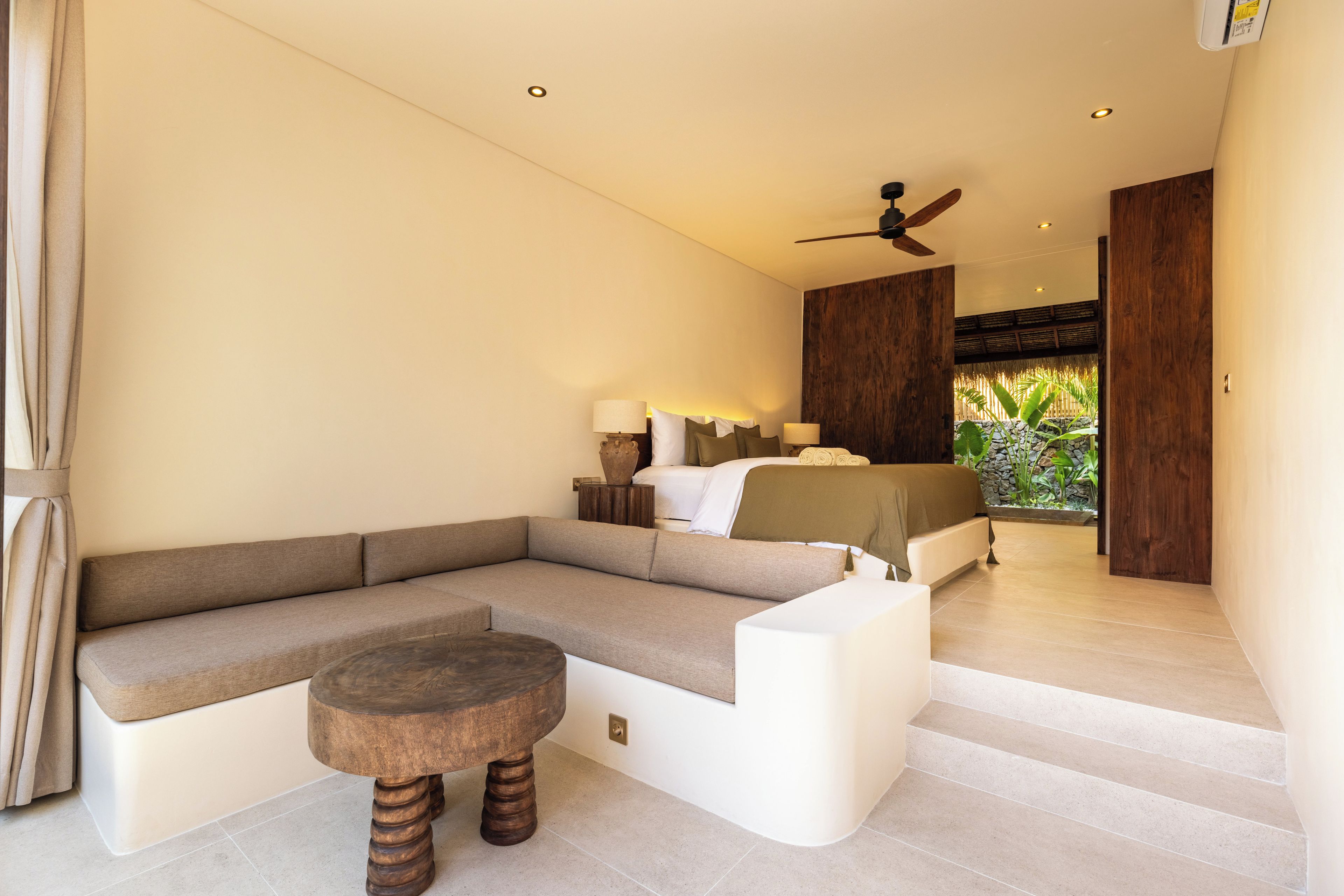 Quarto moderno com sofá, mesa de madeira redonda e cama com vista para o verde.