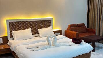 Quarto de hotel com uma cama grande, cabeceira de madeira, mesa de cabeceira, poltrona de couro marrom, pequena mesa e piso de mármore.
