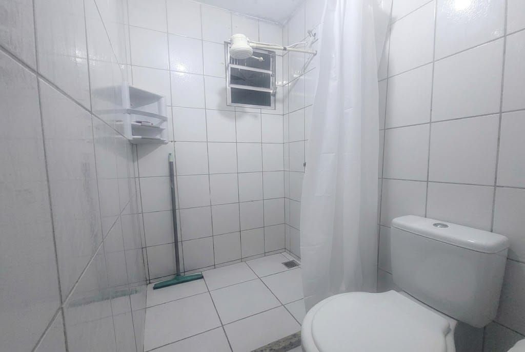 Casa de banho com azulejos brancos, sanita, zona de duche com cortina e uma pequena janela.