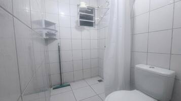 Casa de banho com azulejos brancos, sanita, zona de duche com cortina e uma pequena janela.
