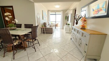 Uma sala de jantar ampla com mesa branca e cadeiras de vime, um sofá e um armário com objetos decorativos.