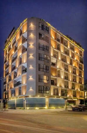 Um hotel de vários andares com varandas, fachada plana e área de estacionamento em frente.