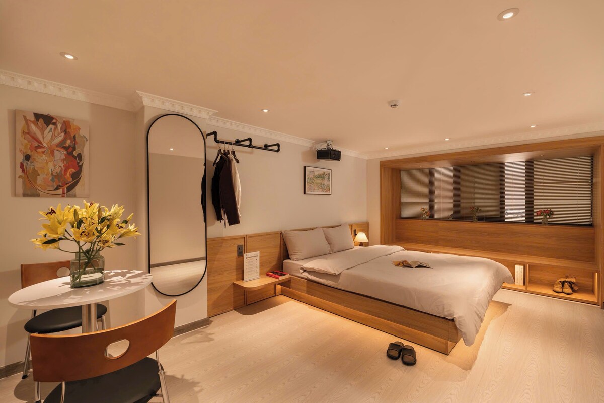 Quarto de hotel moderno com cama, mesa de cabeceira, espelho e vista para o exterior através de grandes janelas.