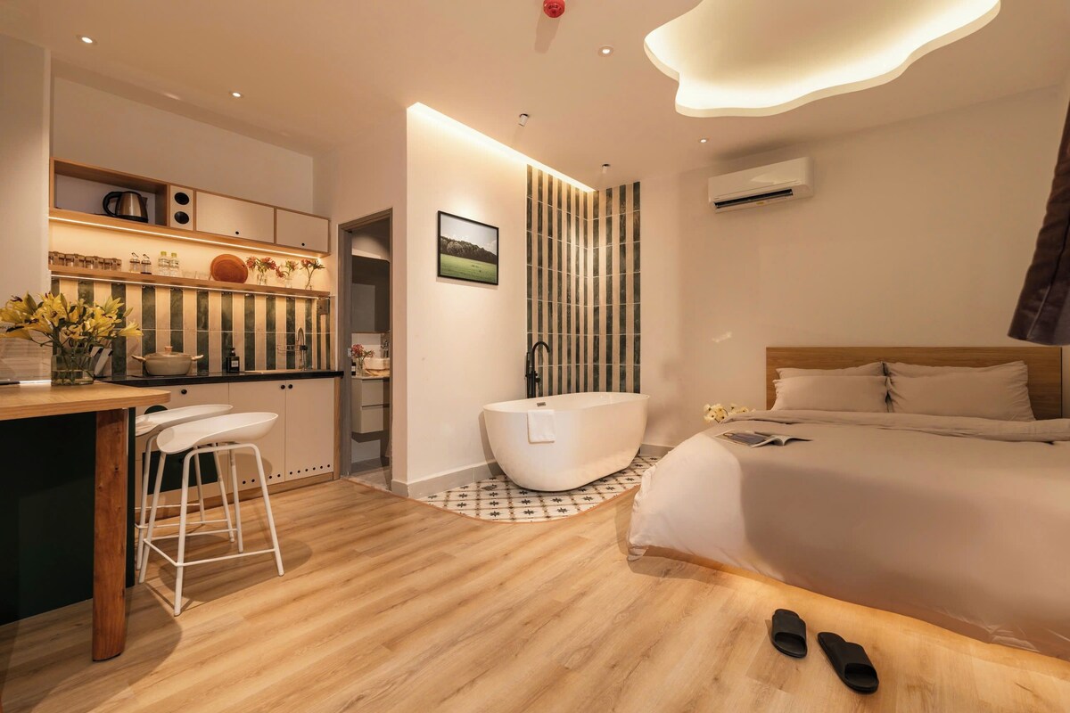 Quarto de hotel moderno com piso de madeira, uma cama grande, banheira, escrivaninha com cadeira e cozinha.