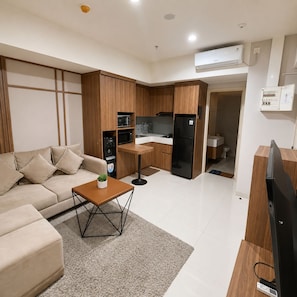 Sala de estar moderna com sofá, mesa de centro e TV. Ao lado, uma cozinha compacta com geladeira, micro-ondas e pia.