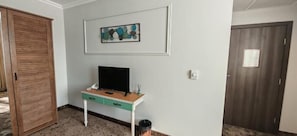 Quarto com guarda-roupa de madeira, secretária com televisão de tela plana, quadro na parede e porta com aviso.