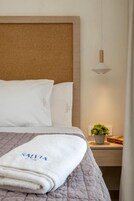 Cama bem arrumada com manta branca e toalha com texto, mesinha de cabeceira com planta e luminária pendente.