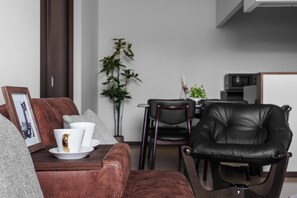 Uma sala de estar moderna com um sofá de couro marrom, uma mesa de centro branca com porta-retrato e xícaras, uma poltrona de couro preta e uma área de jantar com uma planta e uma pequena mesa.
