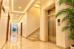 Corredor de hotel com piso de mármore, escada, elevador e planta em vaso.