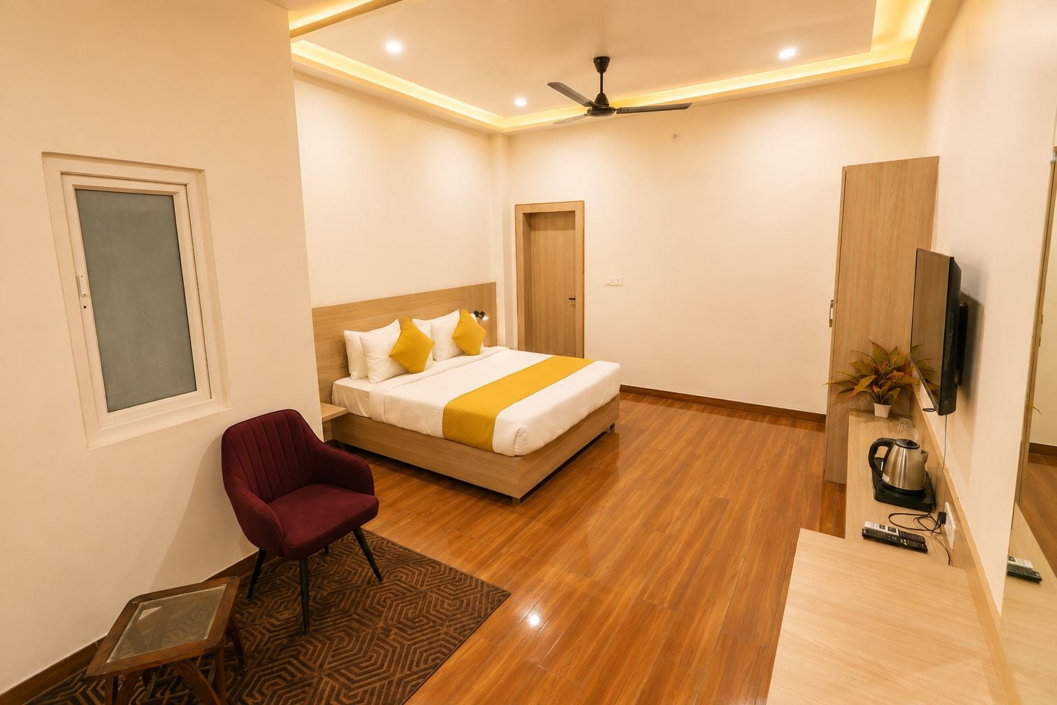 Quarto de hotel com cama, poltrona, televisão, planta e cafeteira.