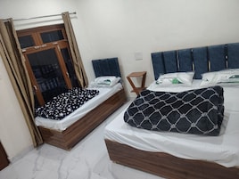 Quarto com duas camas, uma com colcha estampada e outra com manta acolchoada. Há uma janela com cortinas, uma mesinha de cabeceira e uma tomada na parede.